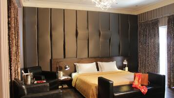 Quarto de hotel moderno com uma cama grande, área de estar com uma poltrona e uma mesa, mesa de cabeceira com abajur, e vista para o exterior pela janela.