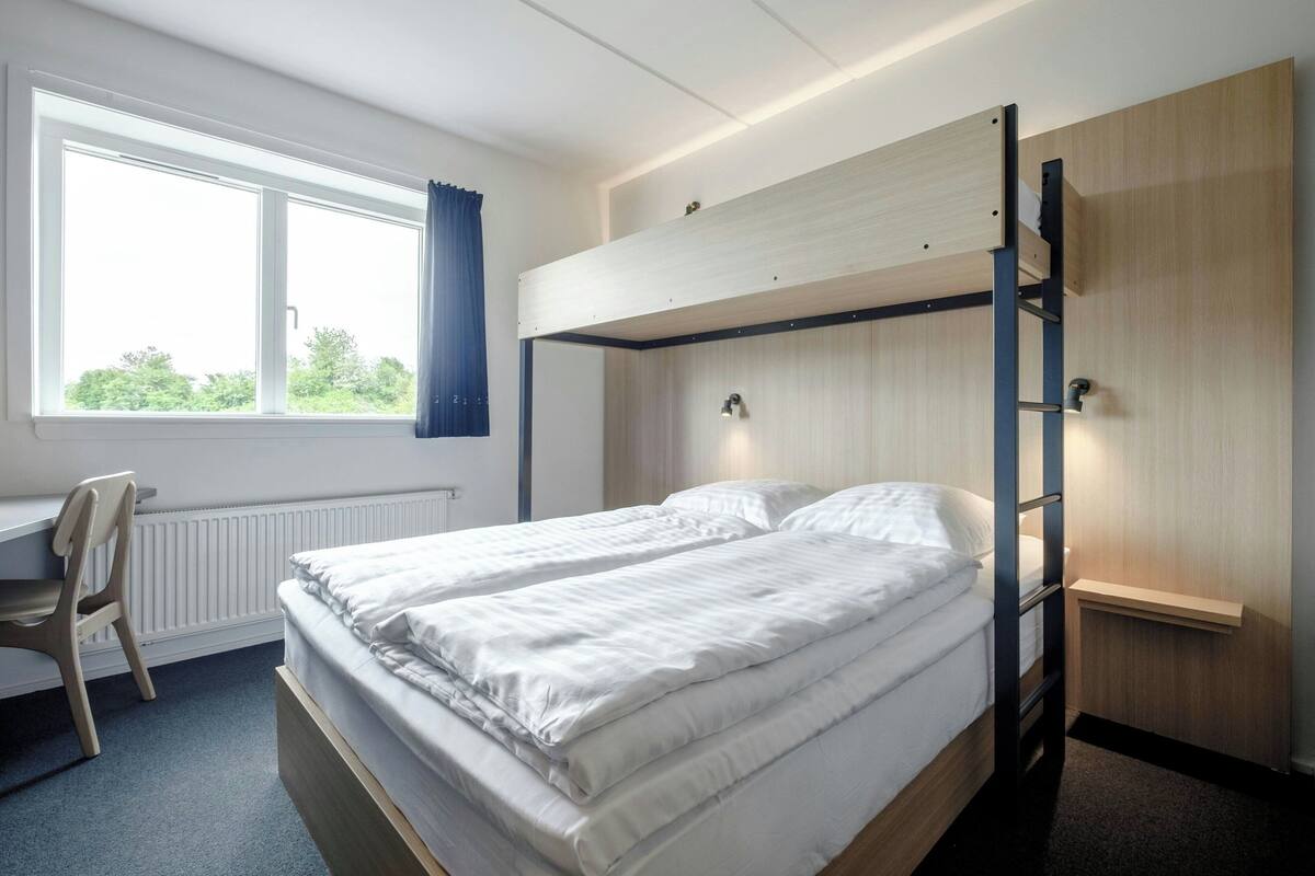 Quarto com beliche, escrivaninha e cadeira, janela com cortinas e parede de madeira.