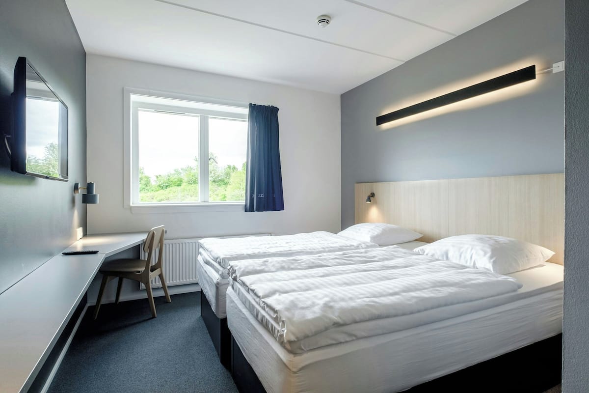 Quarto de hotel com duas camas de solteiro, uma escrivaninha com cadeira, uma janela com cortinas, uma TV fixada na parede e um ponto de luz também fixado na parede.