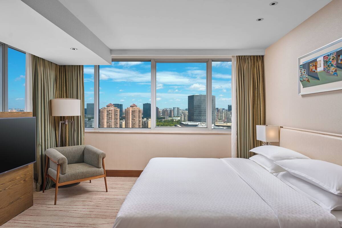 Quarto de hotel com uma cama grande, uma cadeira, uma televisão e vista para o horizonte da cidade através de amplas janelas.