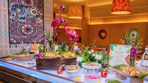Buffet com diversos pratos, tapeçaria decorativa de elefante e flamingos na decoração.