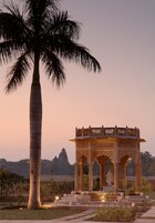 Um pavilhão tradicional com esculturas detalhadas, cercado por um jardim bem cuidado e uma palmeira imponente.