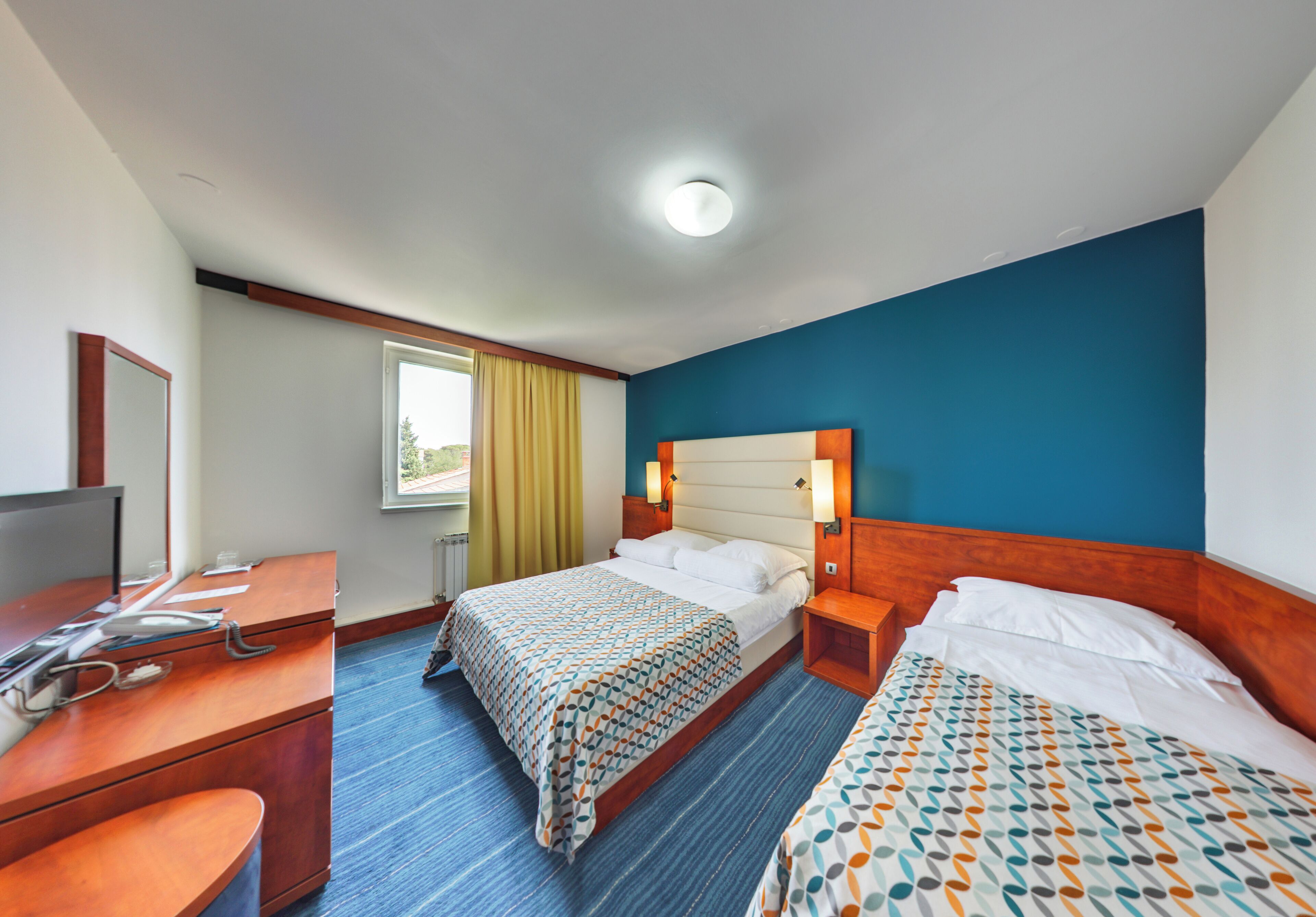 Quarto de hotel com duas camas, uma escrivaninha, uma cadeira, uma janela com cortinas e uma televisão.