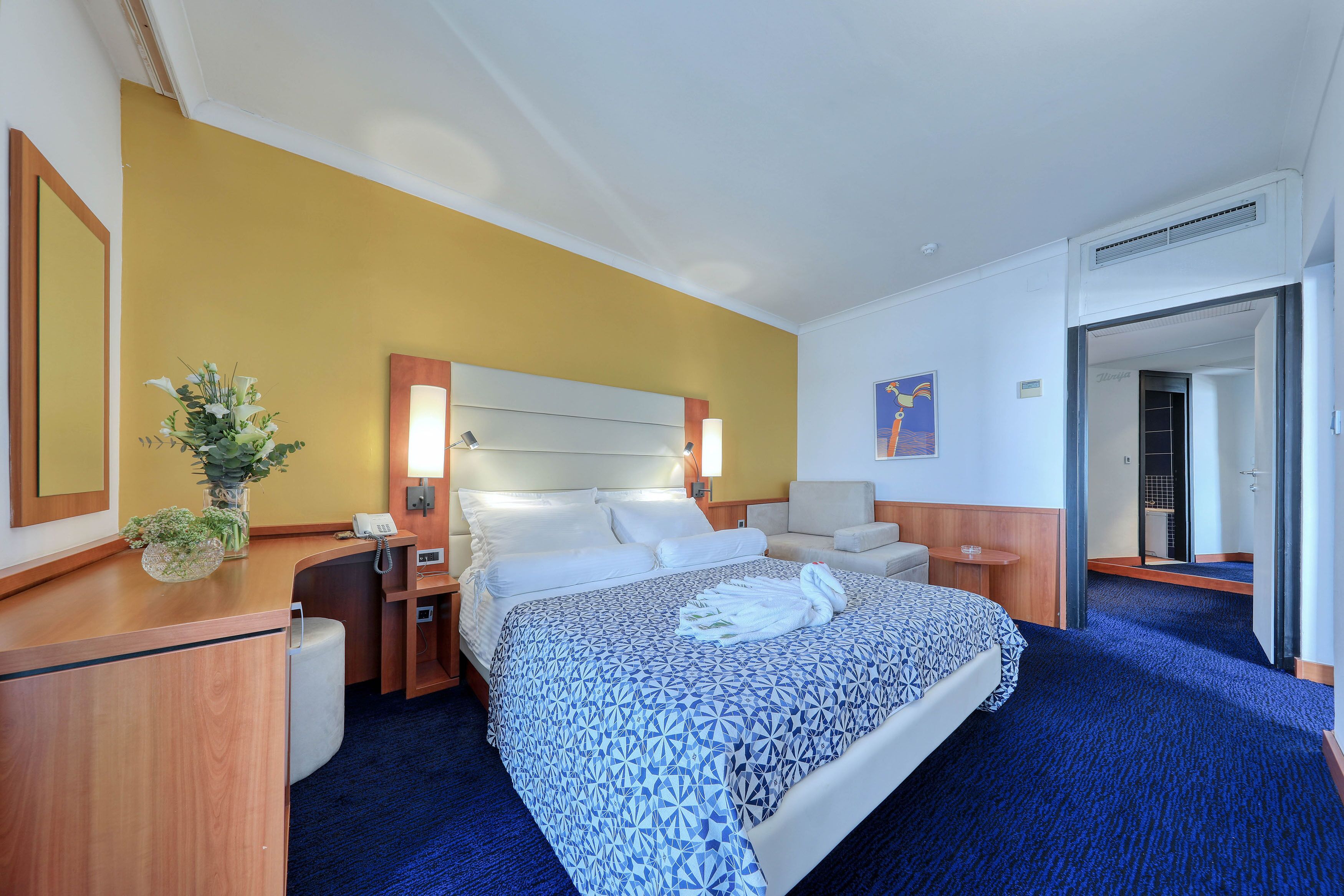 Quarto de hotel com uma cama grande, uma escrivaninha com telefone, um vaso com flores e um espelho na parede.