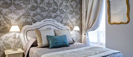 Quarto com papel de parede estampado, cabeceira clássica, duas luminárias de cabeceira e um espelho grande na parede.