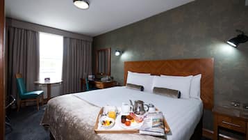Quarto de hotel com cama grande, mesa de cabeceira, escrivaninha com cadeira, janela com cortinas e bandeja com itens de café da manhã.