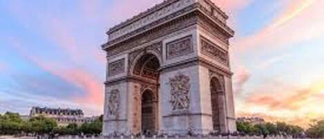 O Arco do Triunfo em Paris, com arquitetura clássica, um arco central e colunas laterais, diante de um céu claro.