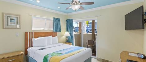 Quarto de hotel com cama, escrivaninha, ventoinha no teto, janela com cortinas e porta para o exterior.