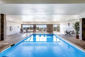 Piscina interior com paredes de vidro transparente, chão revestido de azulejos e área de estar com cadeiras e mesa.