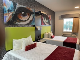 Quarto de hotel com duas camas, cada uma com cabeceira verde, roupa de cama branca e detalhes vermelhos. Há quadros abstratos na parede acima das camas.