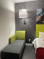 Quarto pequeno e moderno com um sofá cinza, uma poltrona verde, uma cama com cobertor vermelho e travesseiro branco, e uma pintura na parede.
