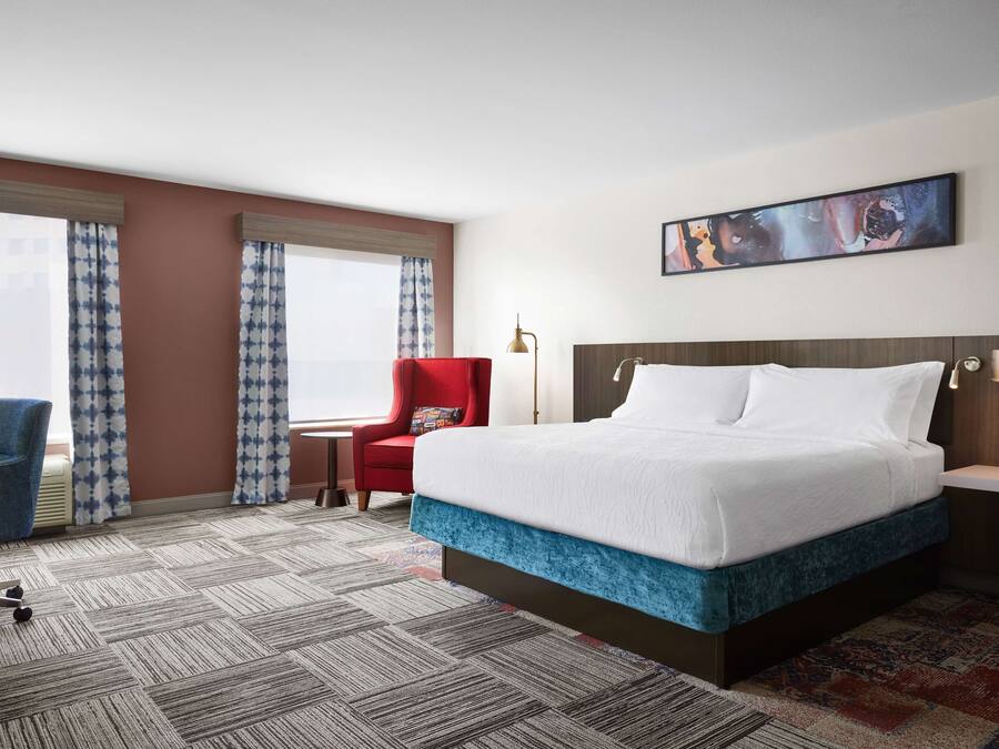 Quarto de hotel com uma cama grande, uma poltrona vermelha, uma cadeira de escritório azul e uma mesa pequena com abajur.
