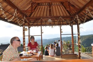 Um gazebo de madeira com telhado de palha, rodeado por mesas e cadeiras, com vista para uma paisagem montanhosa.
