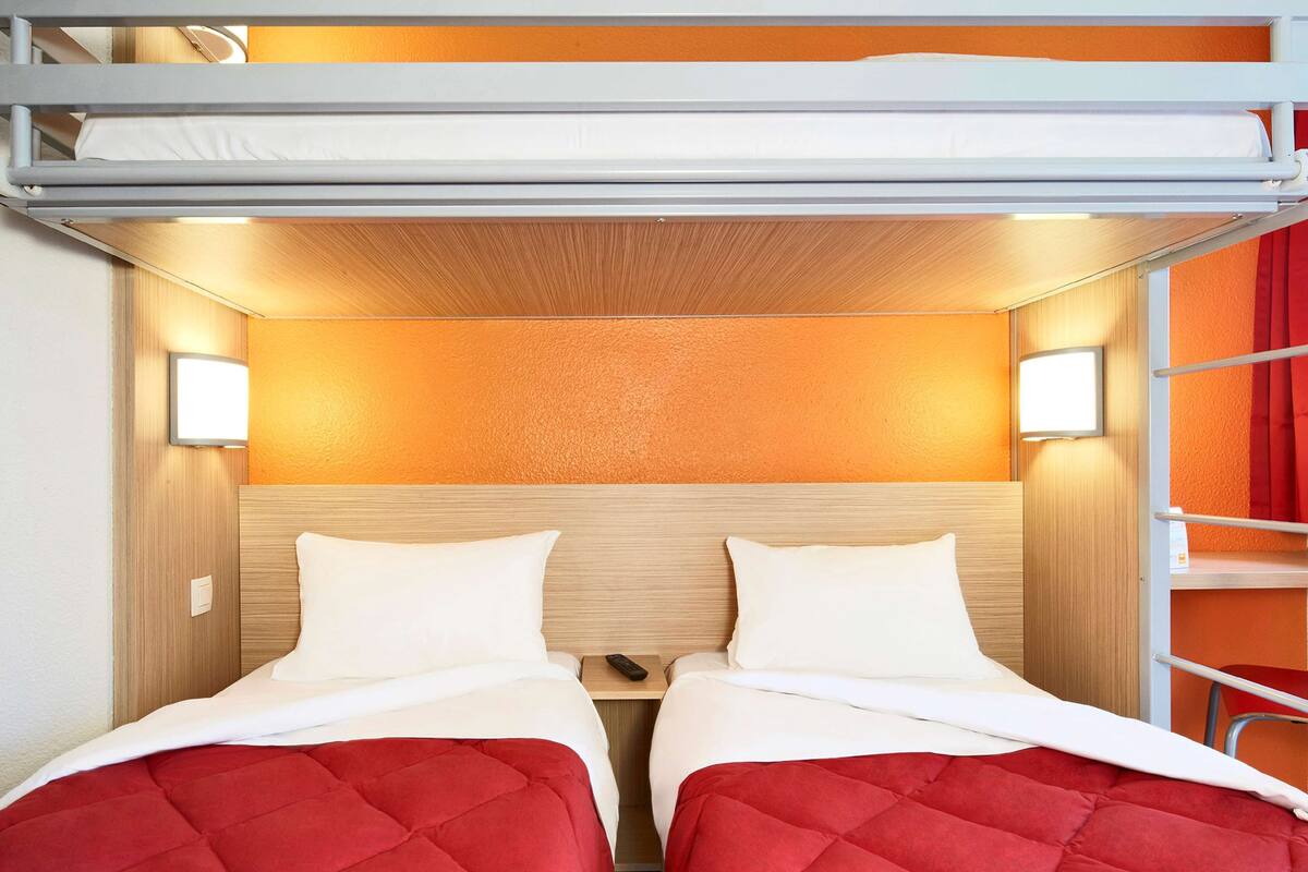 Quarto com beliche, com duas camas, cada uma com cobertura vermelha acolchoada, e cabeceiras de madeira. Existem duas luminárias fixadas na parede, de cada lado das cabeceiras.