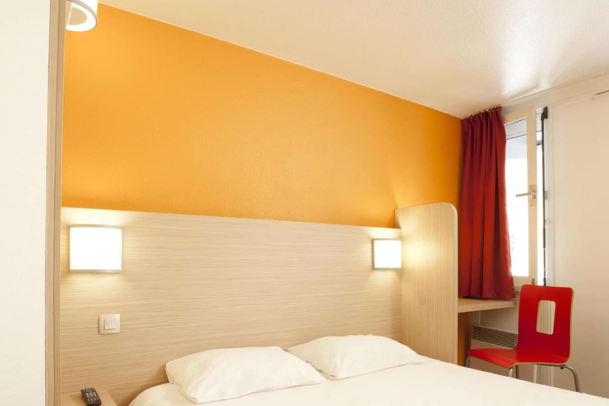 Quarto de hotel com cama, mesa de cabeceira, uma cadeira vermelha e janela com cortinas.