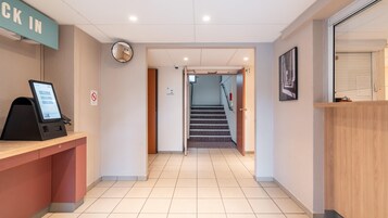 Uma área de recepção de hotel com um quiosque digital, uma placa 'CK IN', uma placa indicando 'proibido fumar' e uma escadaria que leva a outro andar.
