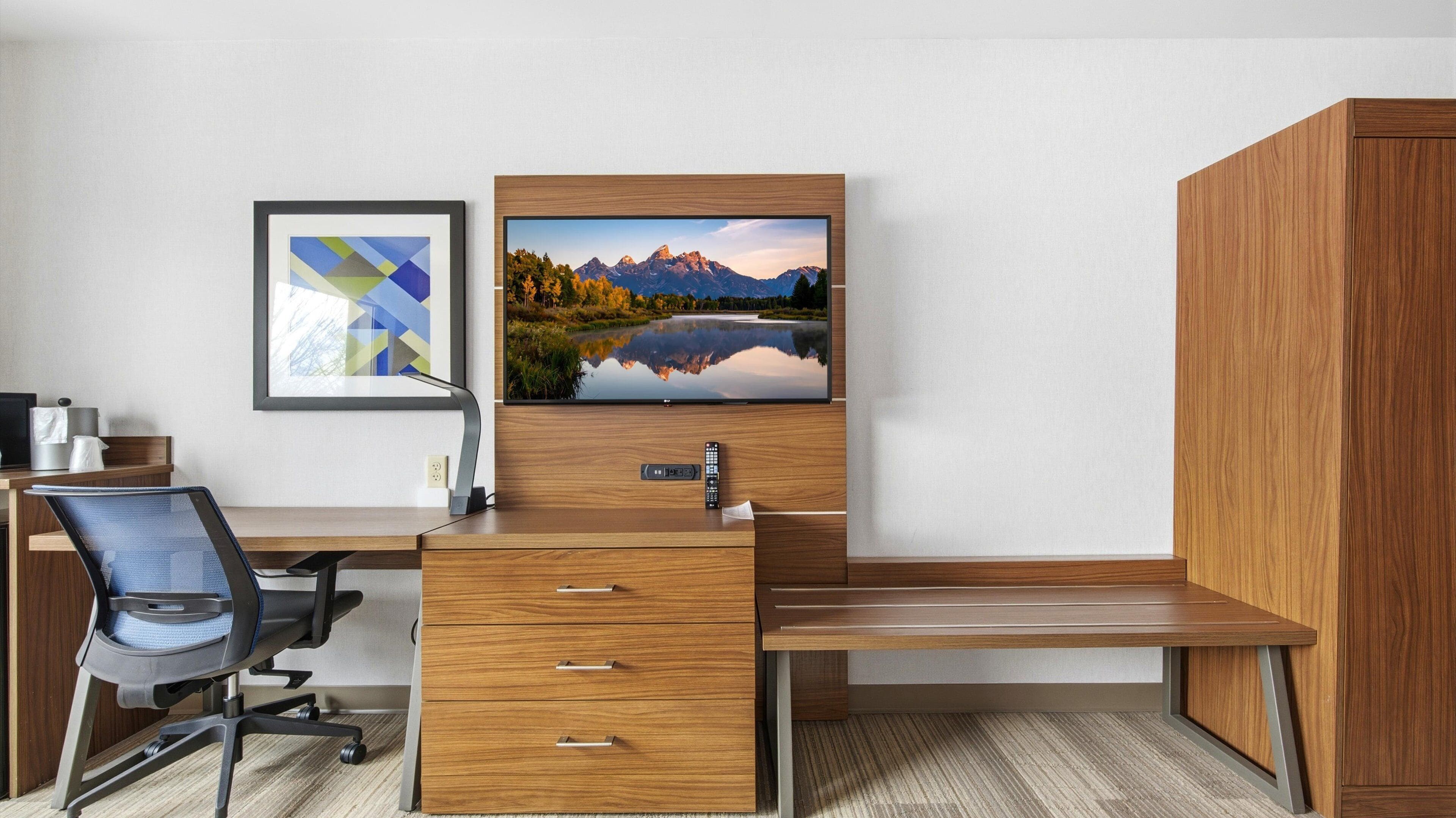 Quarto de hotel com escrivaninha de madeira, uma cadeira, televisão de tela plana na parede e um quadro com paisagem de montanhas.