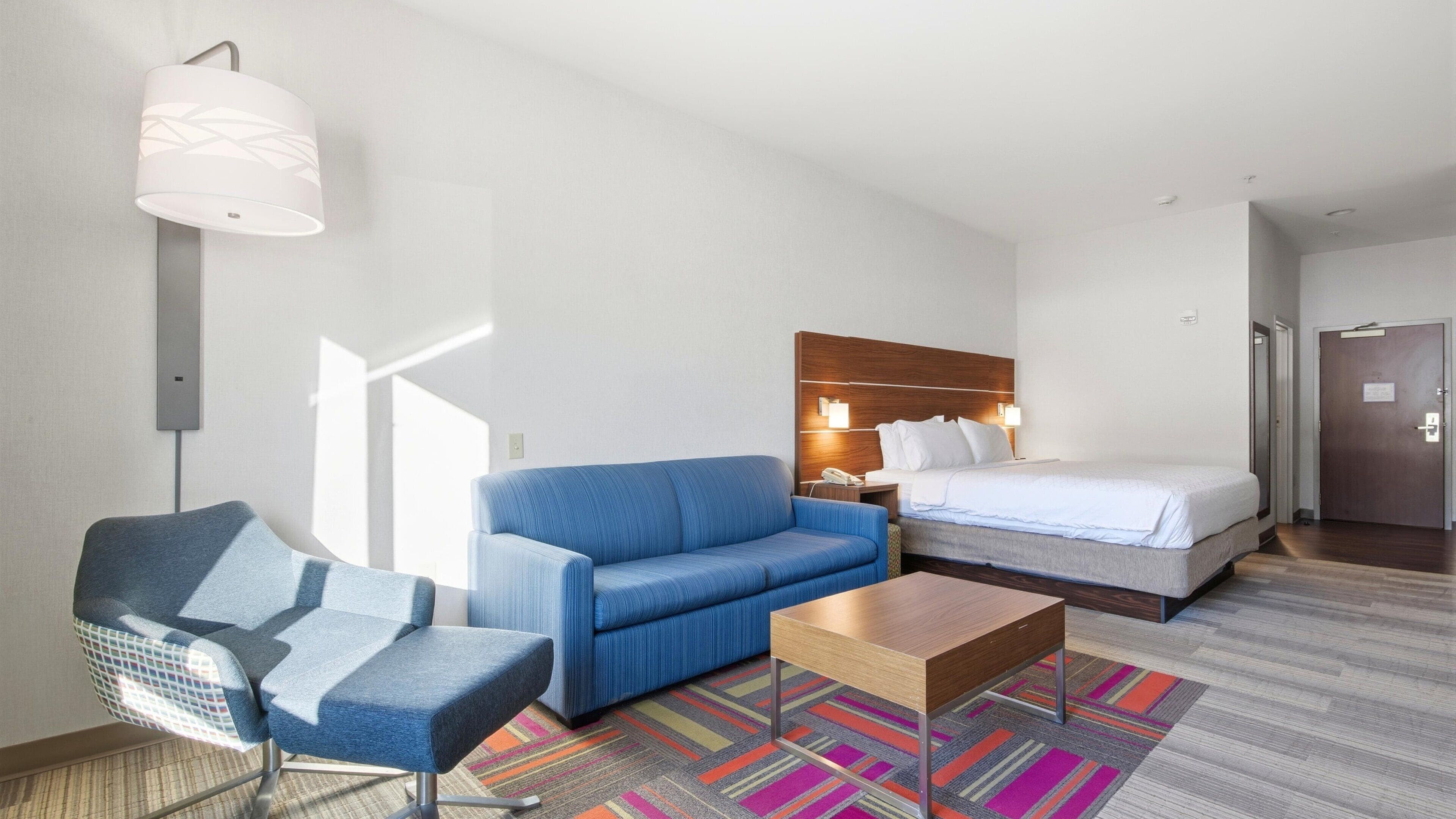 Quarto de hotel com cama, sofá azul, pequena mesa, cadeira e tapete colorido.