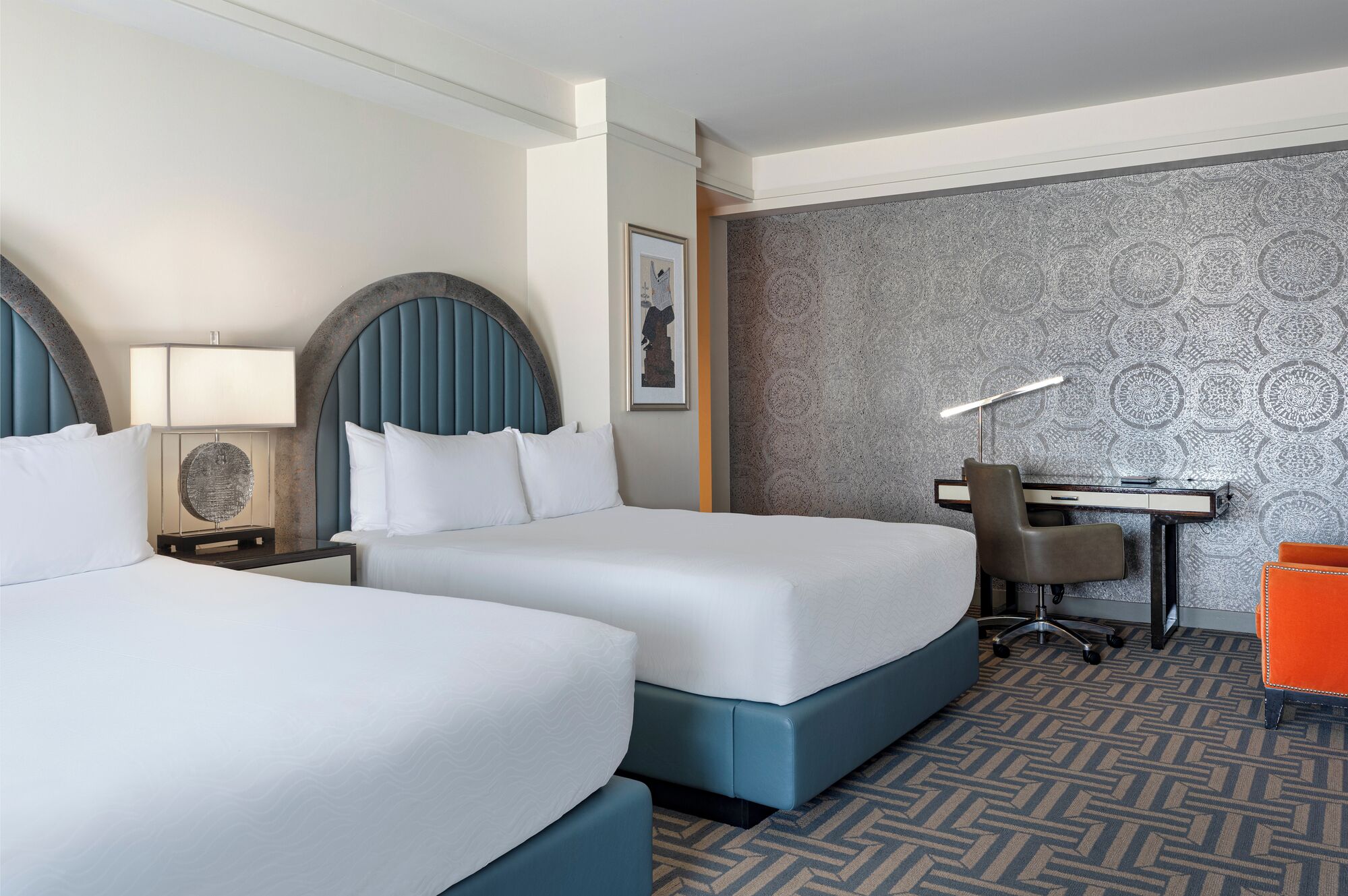 Quarto de hotel com duas camas, uma escrivaninha, uma cadeira e uma luminária.