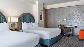 Quarto de hotel com duas camas, uma escrivaninha, uma cadeira e uma luminária.