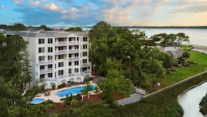 Hotel de vários andares com piscina, rodeado por vegetação exuberante e vista para um lago.