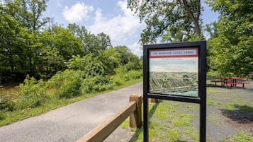 Uma placa com o título 'The Windsor Locks Canal' exibindo um mapa e texto, situada ao lado de um caminho pavimentado em um parque com árvores e área para piquenique.