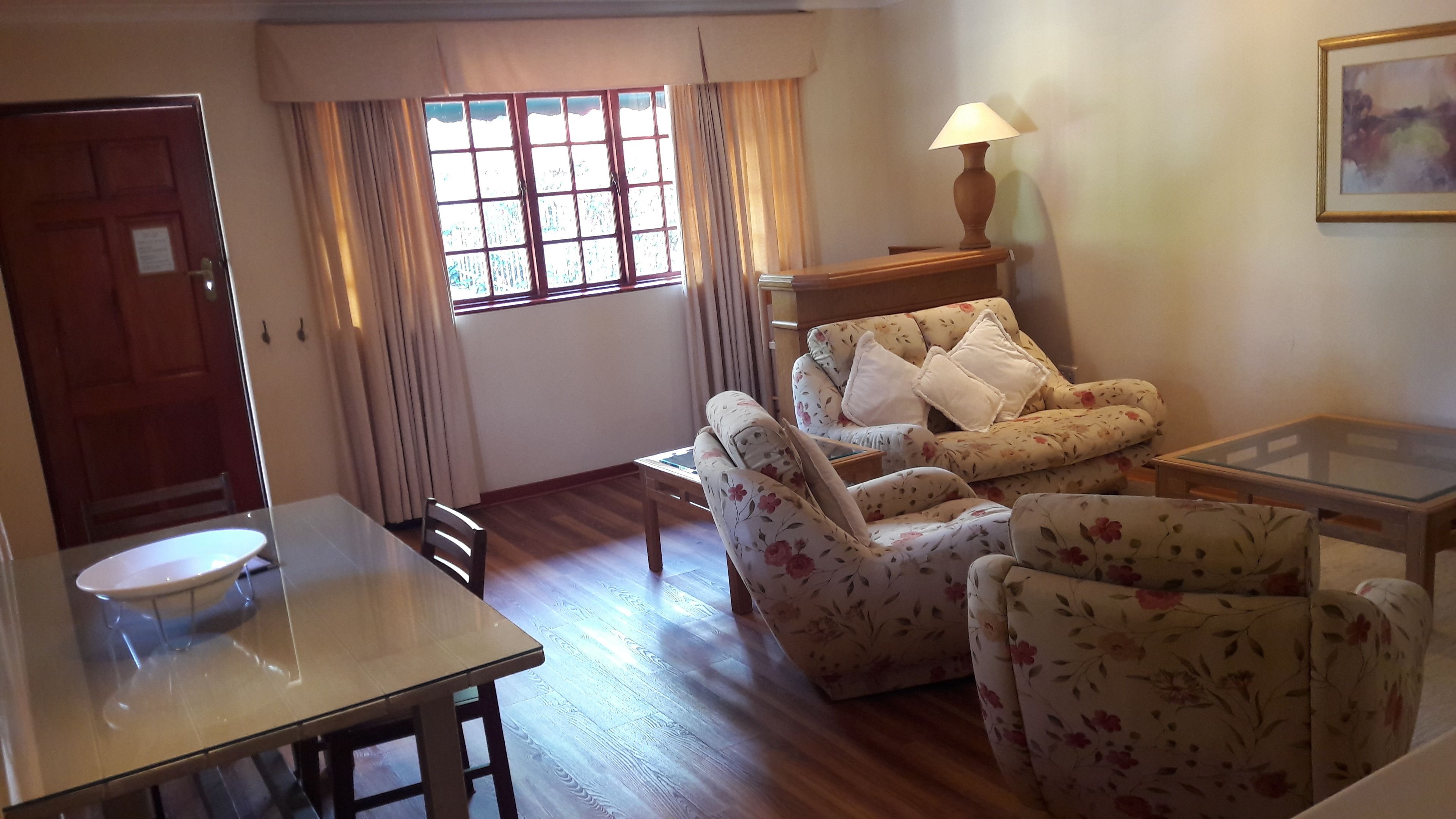 Uma sala de estar com piso de madeira, uma mesa de jantar, duas poltronas florais, uma mesa lateral com abajur e uma janela com cortinas.