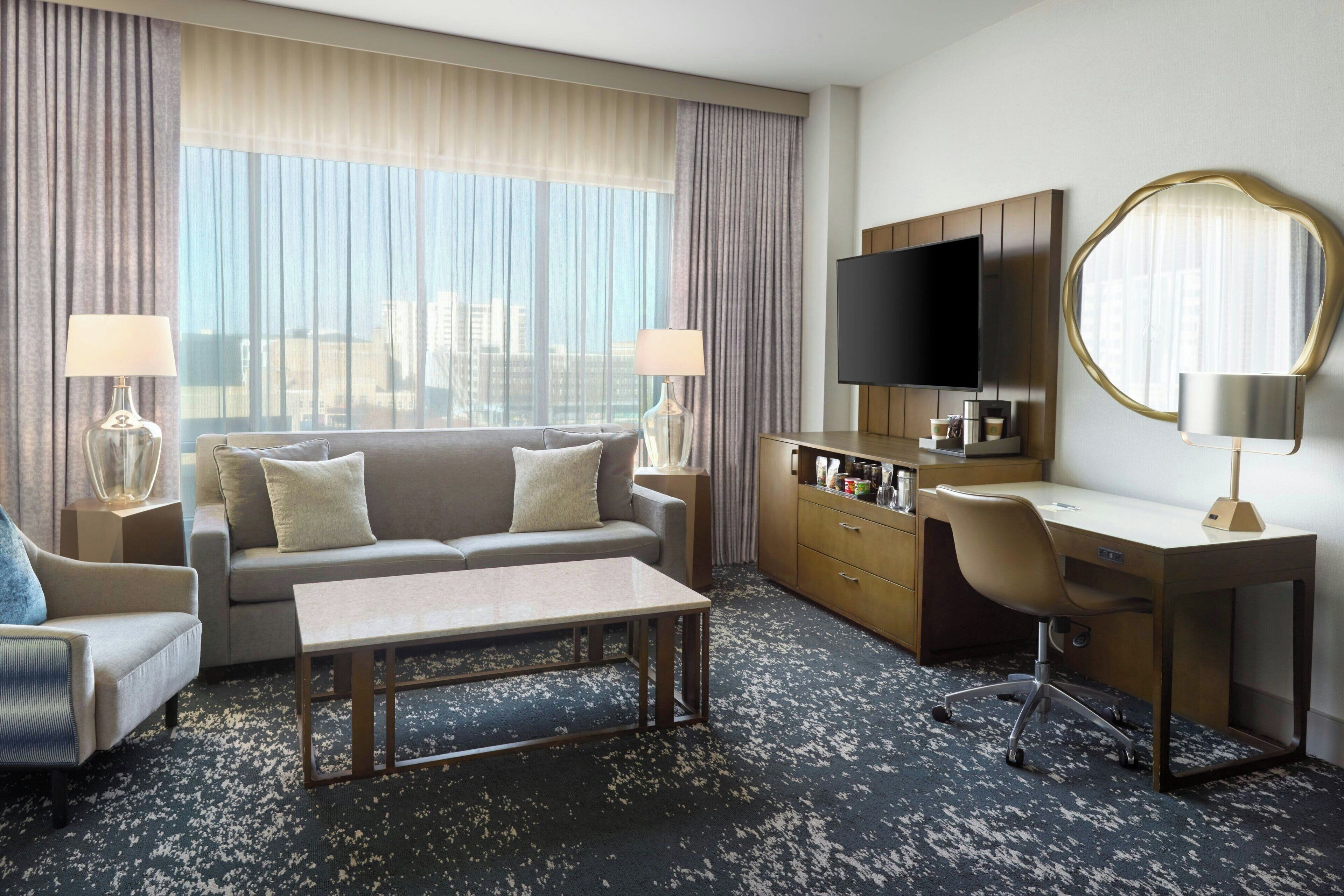 Quarto de hotel com sofá, escrivaninha, televisão e uma janela grande com cortinas.