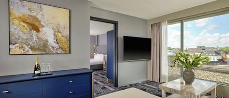 Quarto de hotel moderno com uma grande pintura abstrata, minibar, TV de tela plana, uma ampla janela com vista para a cidade e uma pequena mesa com um vaso de flores.