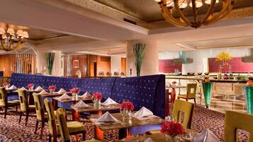 Um restaurante com um assento acolchoado azul, mesas postas com guardanapos brancos e flores cor-de-rosa, e uma área de bar ao fundo.