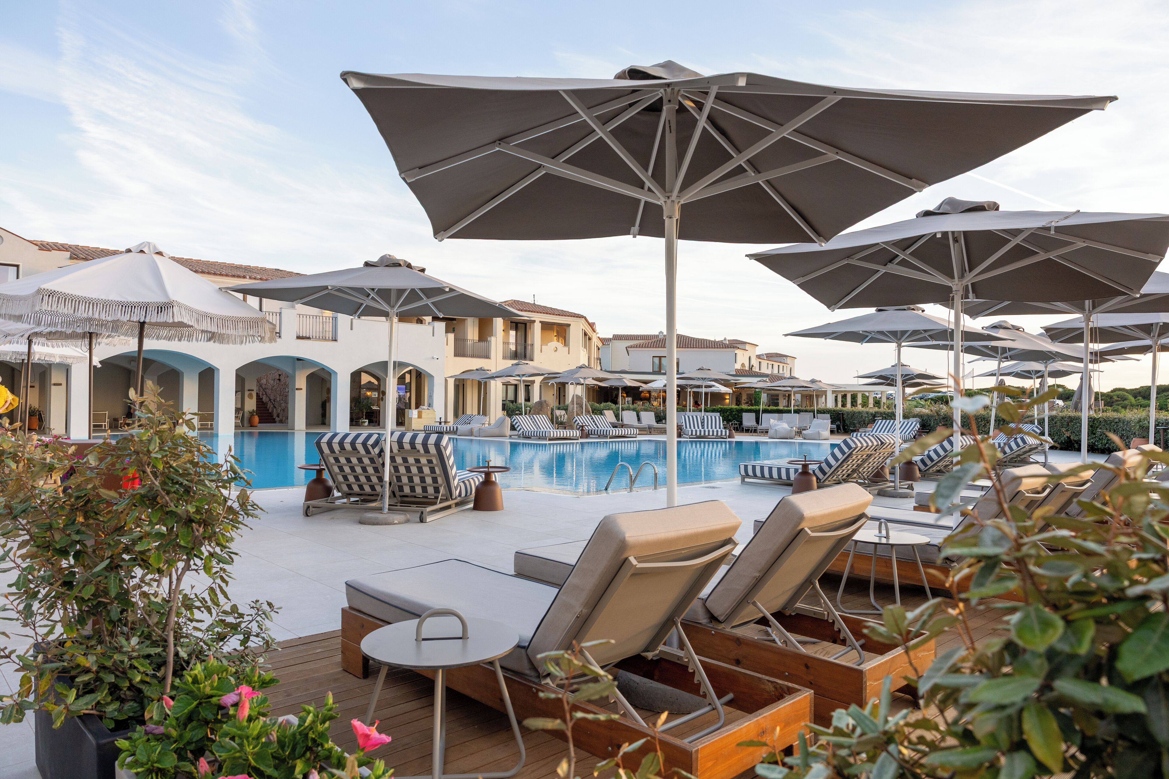 Uma área à beira da piscina com espreguiçadeiras e guarda-sóis, uma piscina e edifícios ao fundo.