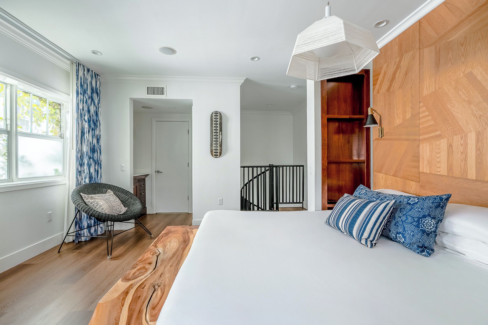 Quarto com cama, cadeira, cabeceira de madeira, luminária pendente e vista para uma escada.