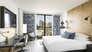 Quarto de hotel moderno com uma cama grande, uma escrivaninha com luminária, uma cadeira e vista para a cidade pela janela.