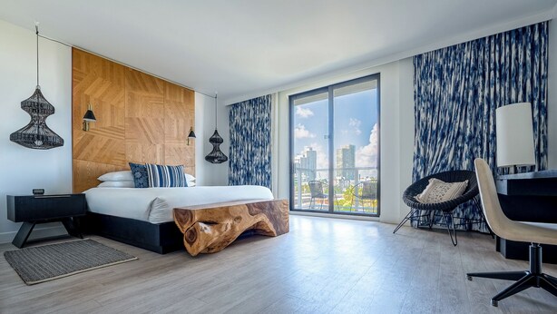 Quarto moderno com uma cama grande, cabeceira de madeira, vista para a cidade e varanda com cadeiras.