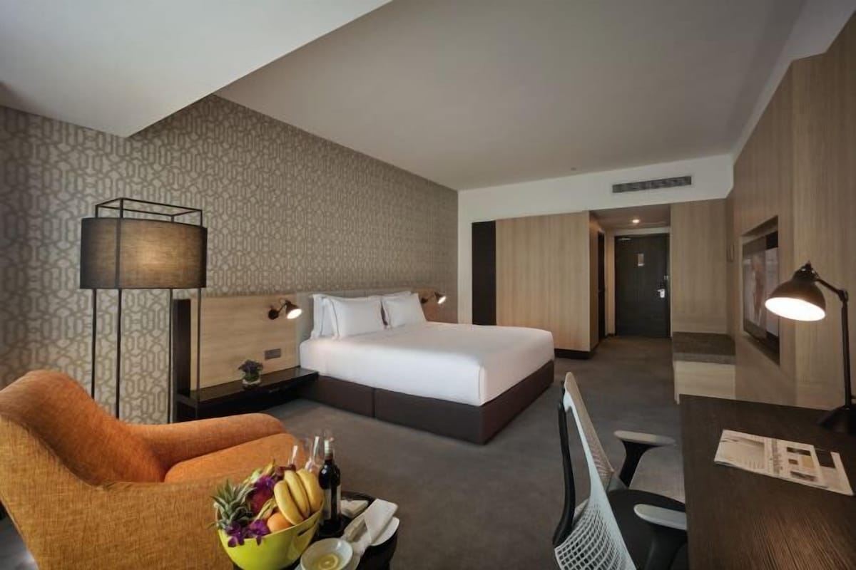 Quarto de hotel com uma cama grande, uma escrivaninha com cadeira, uma luminária e uma mesinha com uma fruteira e bebidas.
