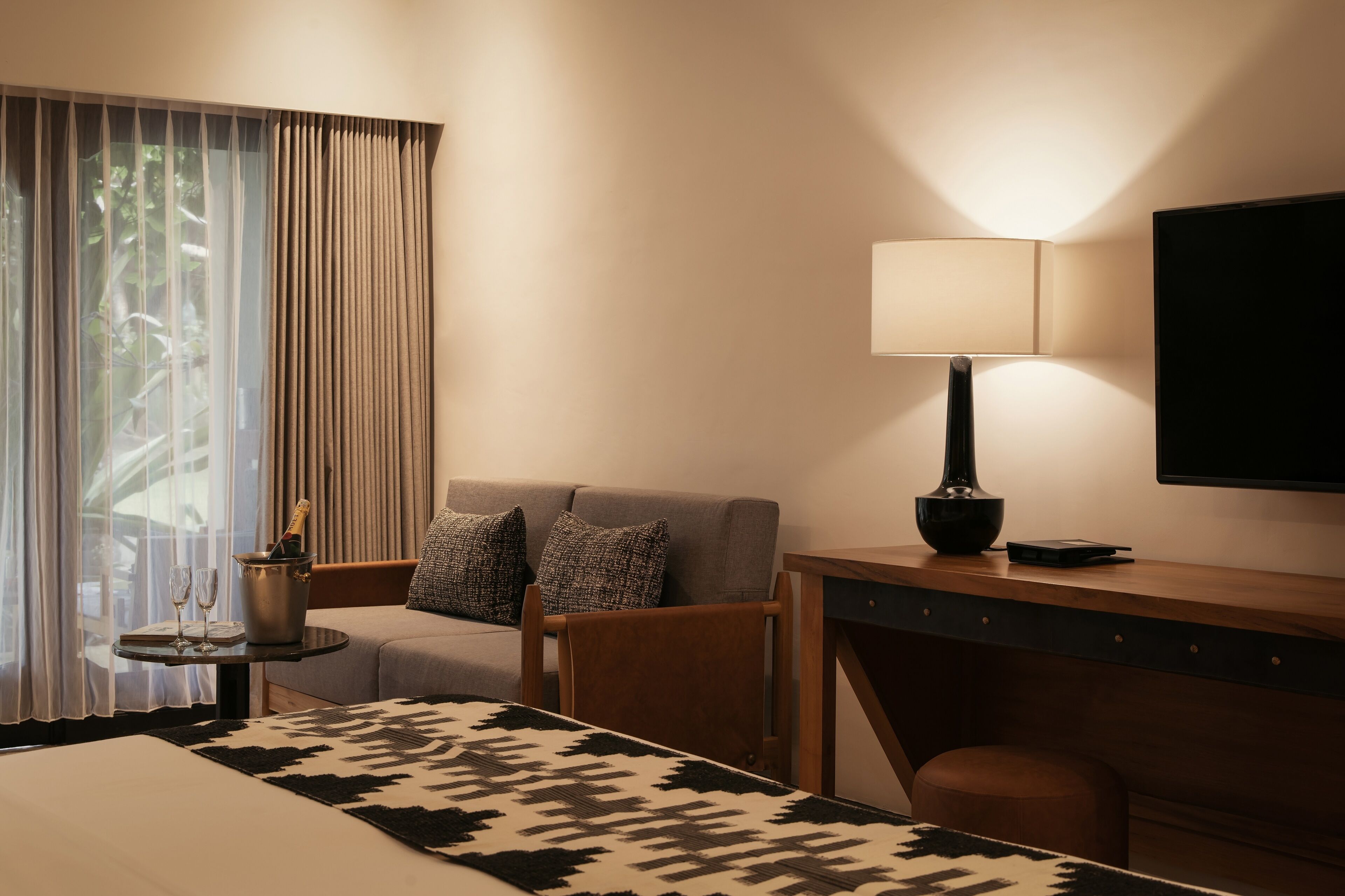 Quarto de hotel com cama, sofá, escrivaninha, luminária, televisão e janela com cortinas.