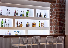 Uma área de bar com várias prateleiras de garrafas de bebidas, uma parede de tijolos e quatro cadeiras brancas.