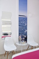 Quarto com cama, duas cadeiras brancas, uma mesa redonda pequena com um livro e um espelho na parede refletindo uma vista panorâmica da água.