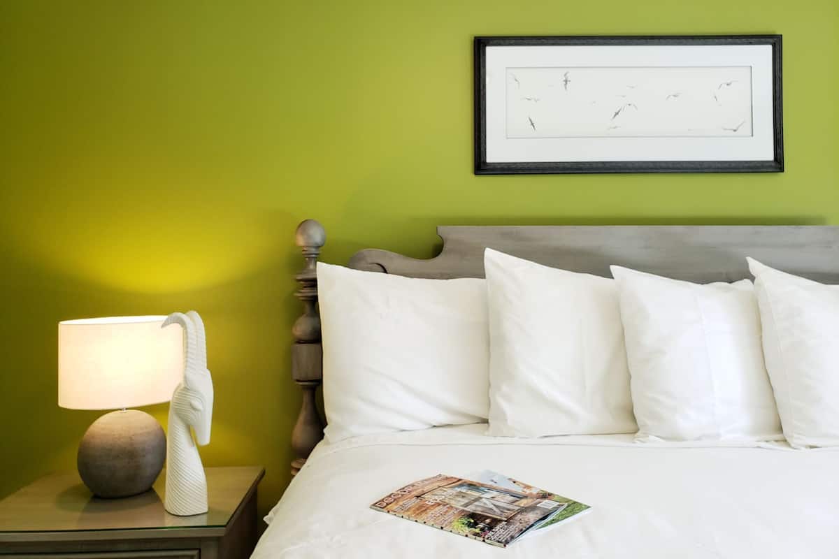 Quarto de hotel com cama, mesa de cabeceira com abajur, quadro na parede e revista sobre a cama.