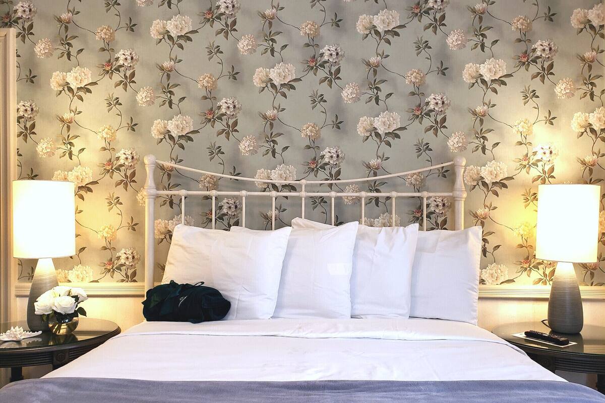 Quarto de hotel com papel de parede floral, estrutura de cama de metal branco, roupa de cama branca e duas luminárias de cabeceira.