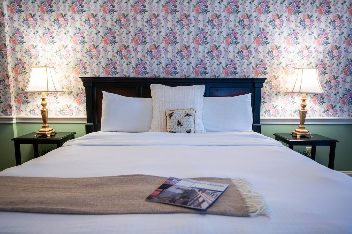 Cama bem arrumada com lençóis brancos, um travesseiro decorativo e uma revista sobre um cobertor. Há duas luminárias de cabeceira em mesas de cabeceira combinando.