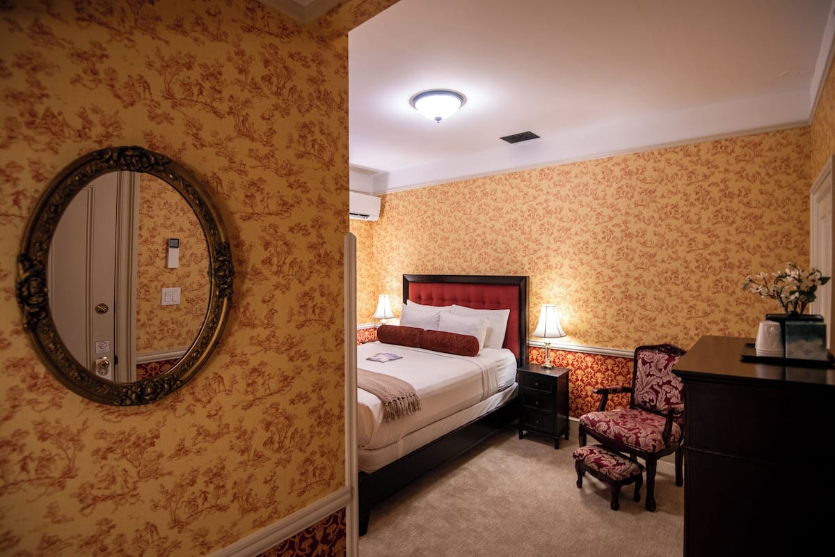 Quarto de hotel com papel de parede floral, um espelho grande e ricamente decorado, cama com lençóis brancos, cabeceira vermelha, mesa de cabeceira com abajur, uma cadeira e um armário.