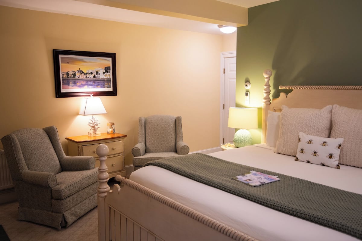 Quarto de hotel com uma cama, duas poltronas, uma mesinha de cabeceira e um quadro na parede.