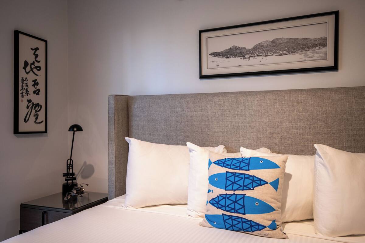 Cama bem arrumada com almofadas brancas e azuis, um quadro emoldurado acima e uma pequena mesa com abajur ao lado da cama.