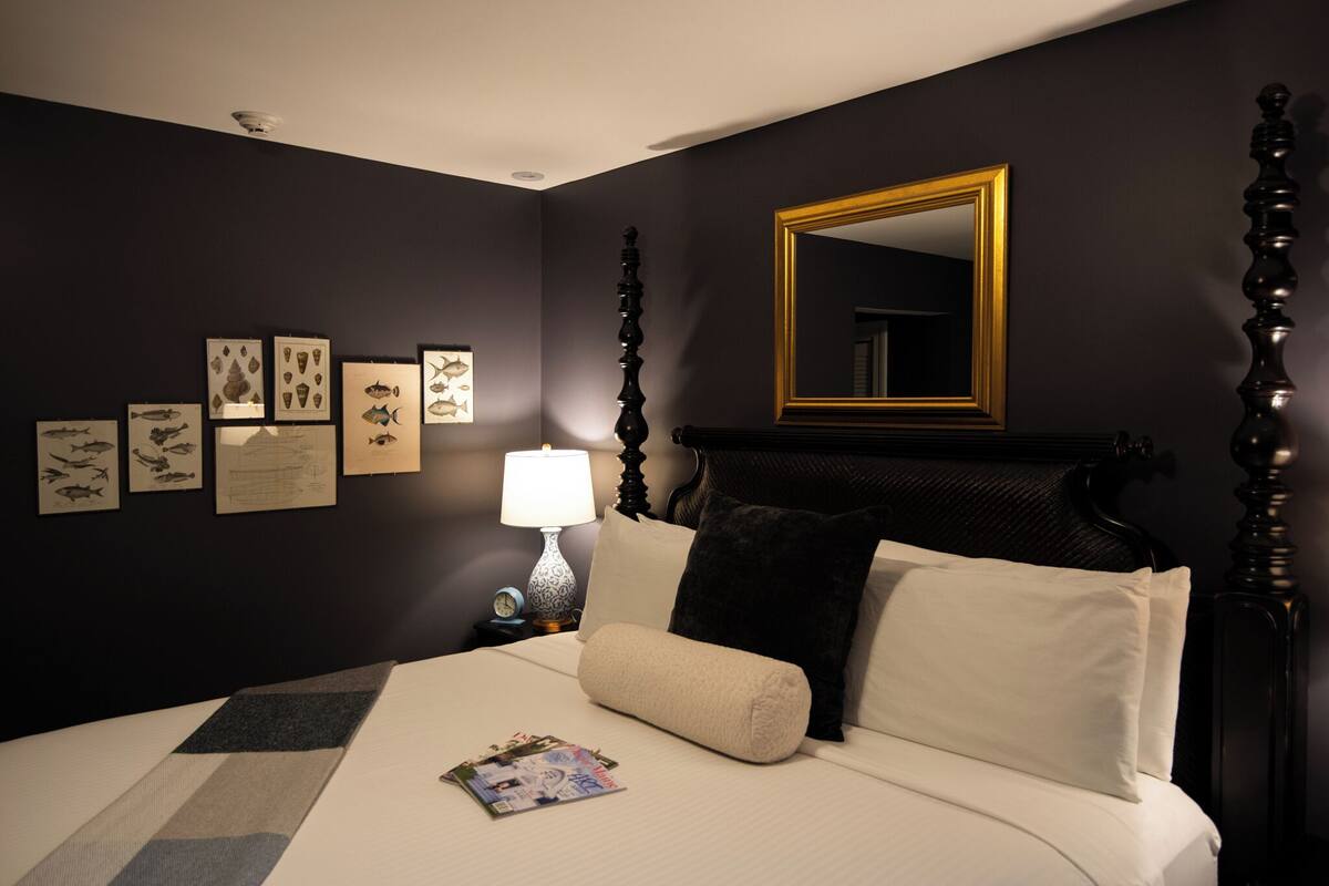 Um quarto com cama, mesa de cabeceira, abajur, espelho e obras de arte nas paredes.