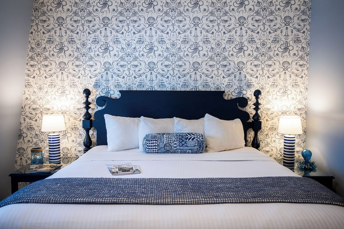 Cama bem arrumada com almofada estampada, uma revista e tapete texturizado.