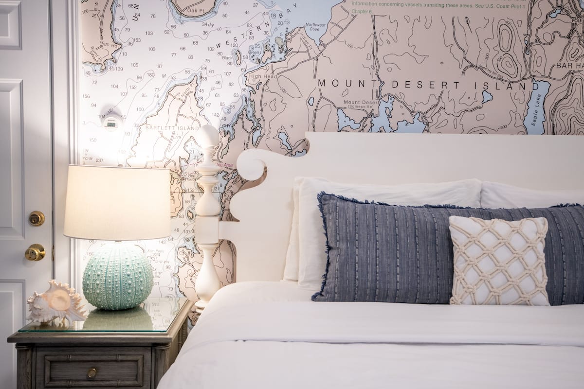 Quarto com parede de mapa, cama com almofadas azuis e bege, abajur e concha decorativa na mesa de cabeceira.