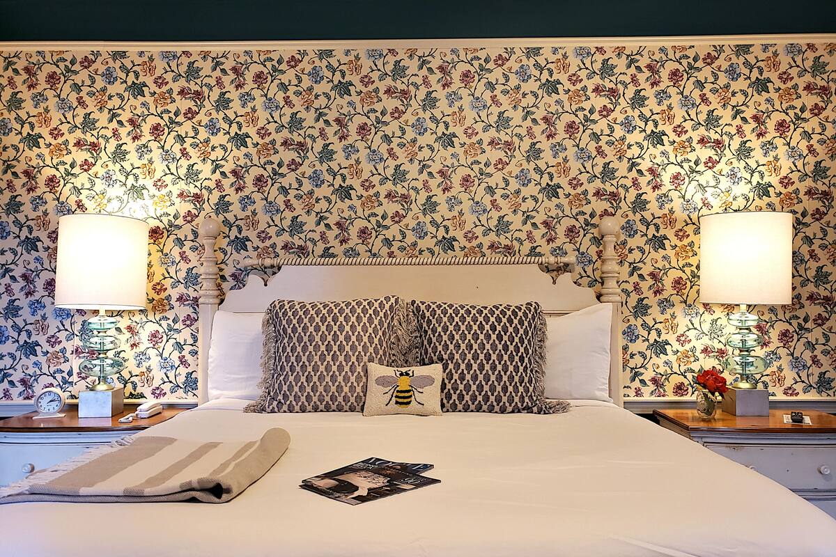 Cama bem arrumada com manta listrada, almofada com desenho de abelha e uma revista sobre a cama. Duas mesas de cabeceira, com abajures e um relógio em uma delas. O quarto tem papel de parede floral e cabeceira com padrão decorativo.