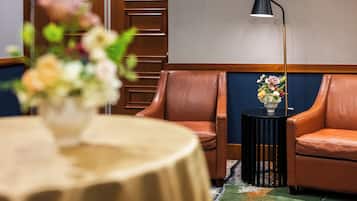 Um quarto com uma mesa redonda, duas poltronas de couro, uma mesinha lateral com um vaso de flores, um abajur e um painel de parede de madeira.
