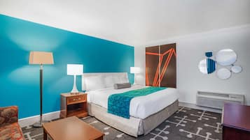 Quarto de hotel com uma cama grande, duas mesas de cabeceira com luminárias, uma pequena mesa de madeira, um tapete estampado e um painel com espelhos decorativos.